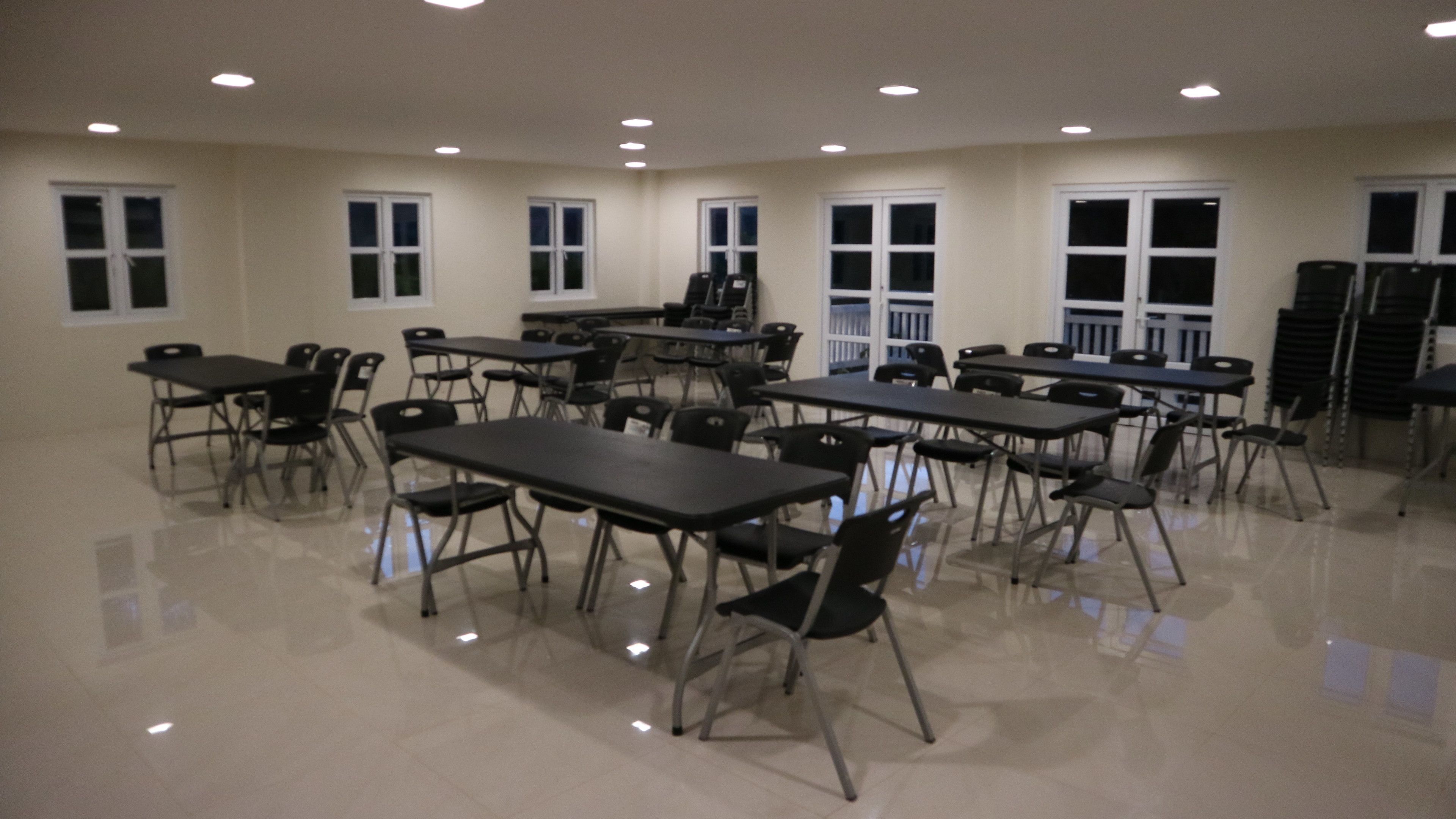 Uma sala de aula com mesas e cadeiras dispostas em fileiras, amplas janelas e luminárias no teto.
