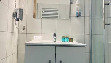 Um banheiro com toucador branco, espelho, secador de mãos, secador de cabelo, box de vidro para chuveiro e lixeira.