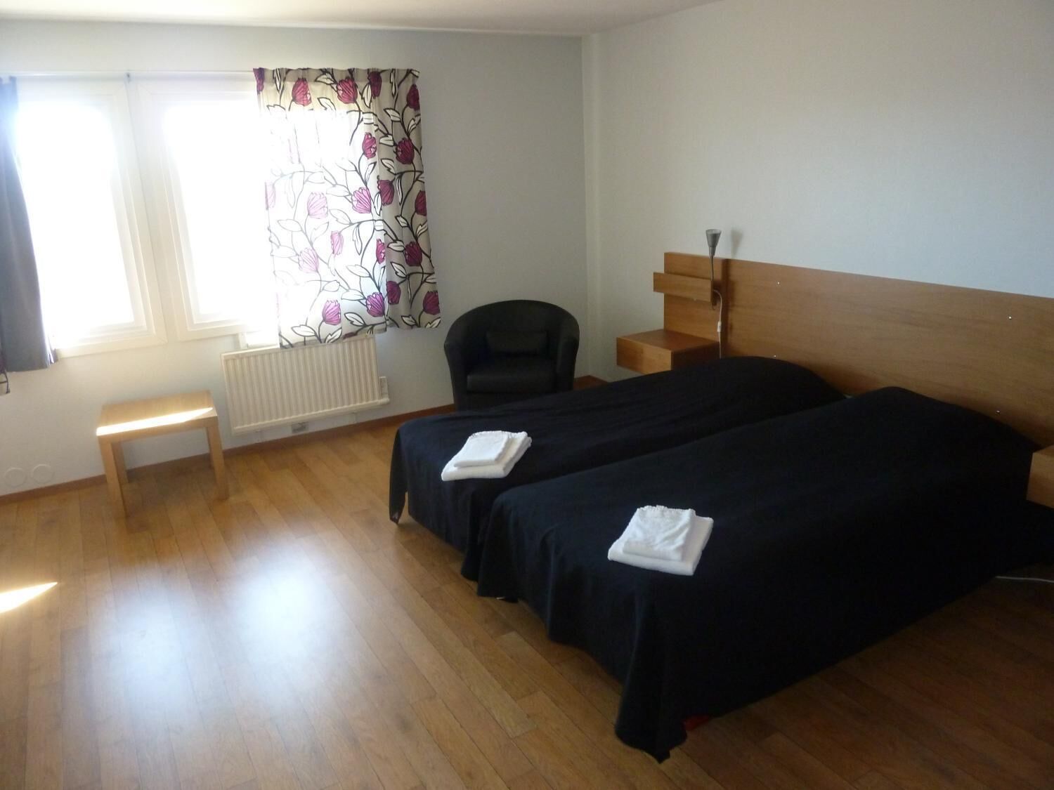 Quarto com duas camas, um banco, uma cadeira, um radiador e uma janela com cortinas.