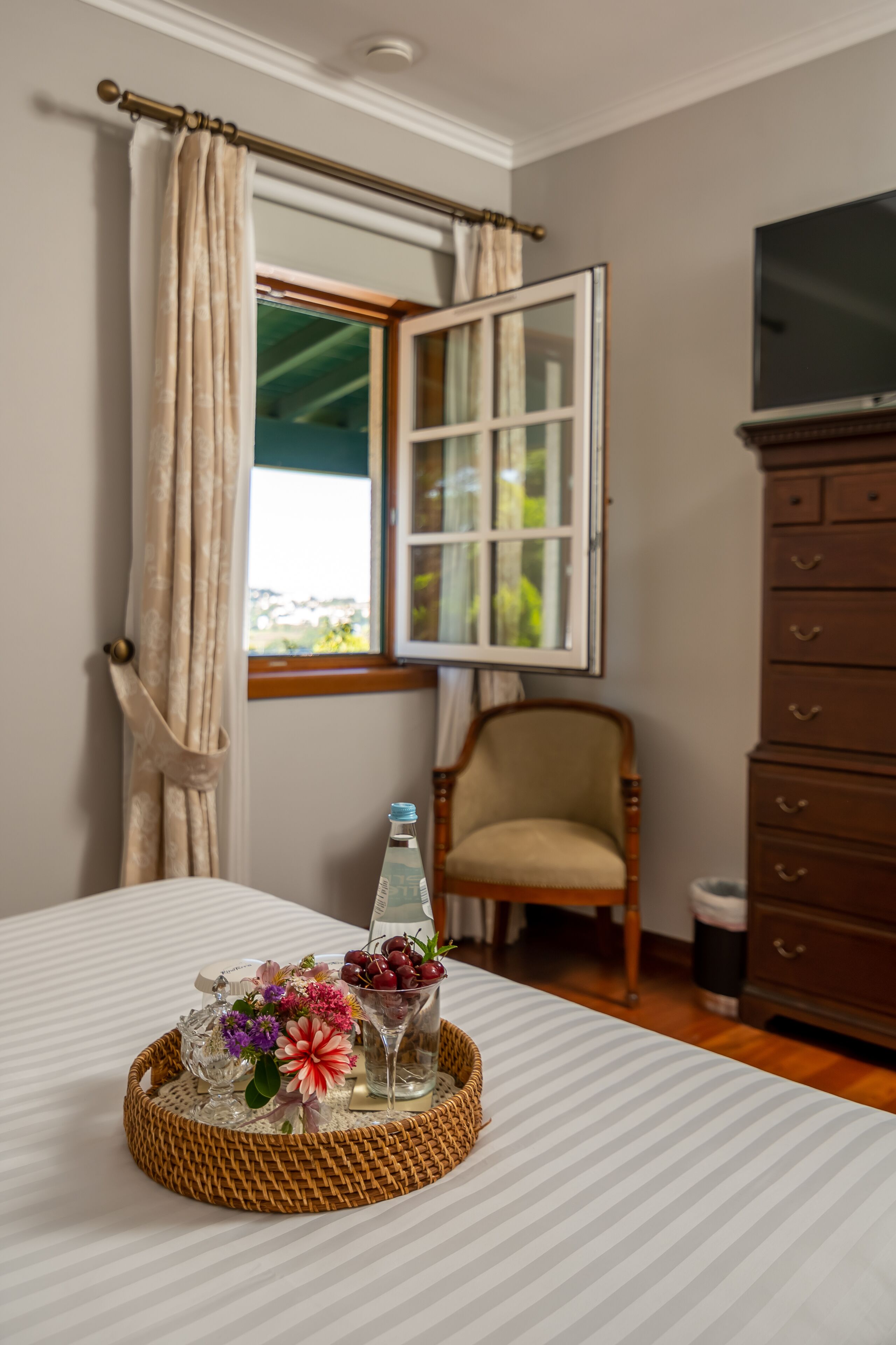 Quarto com cama, bandeja de vime com flores e uma garrafa, cadeira, cômoda e janela com cortinas.