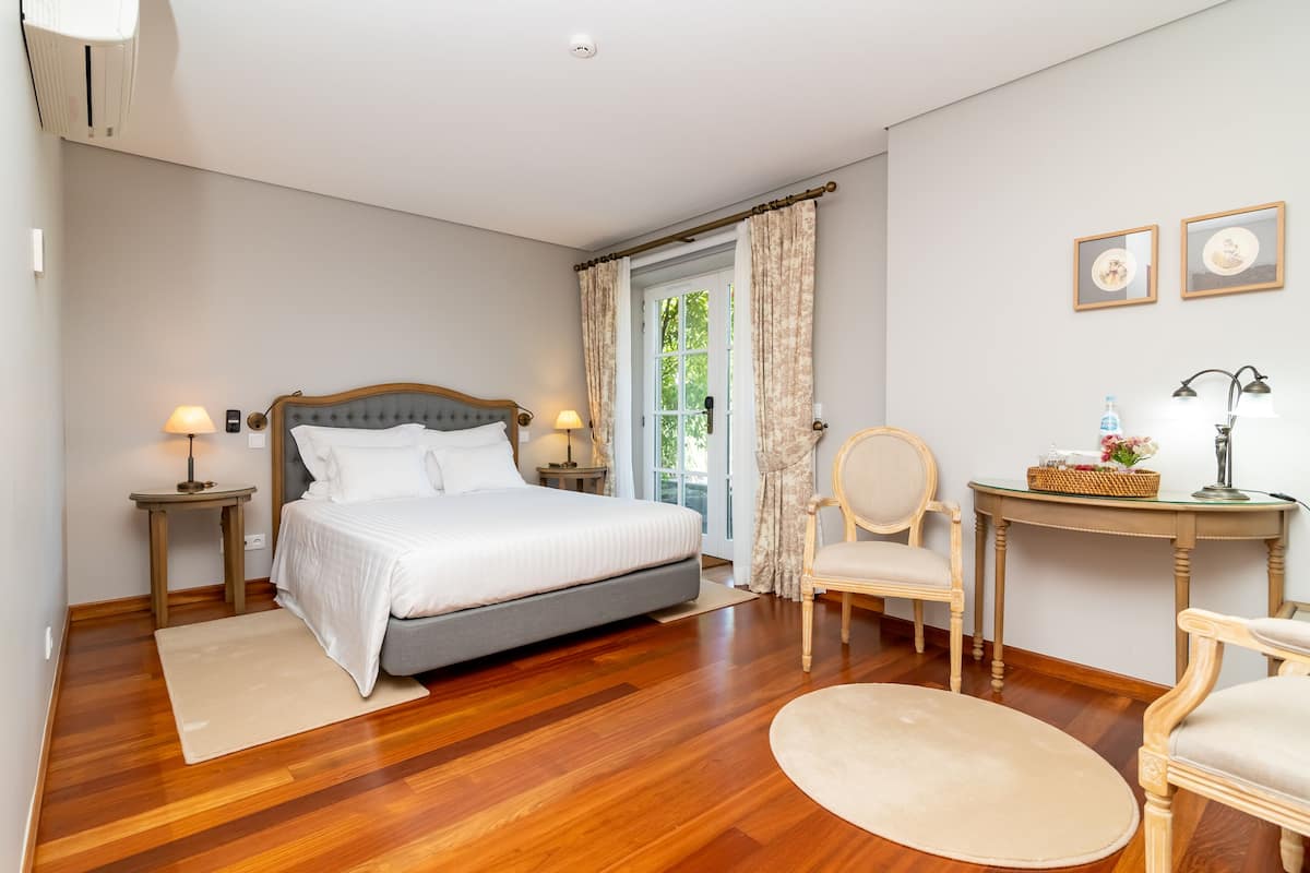 Quarto com cama, mesas de cabeceira, uma cadeira, uma mesinha e uma janela com cortinas.