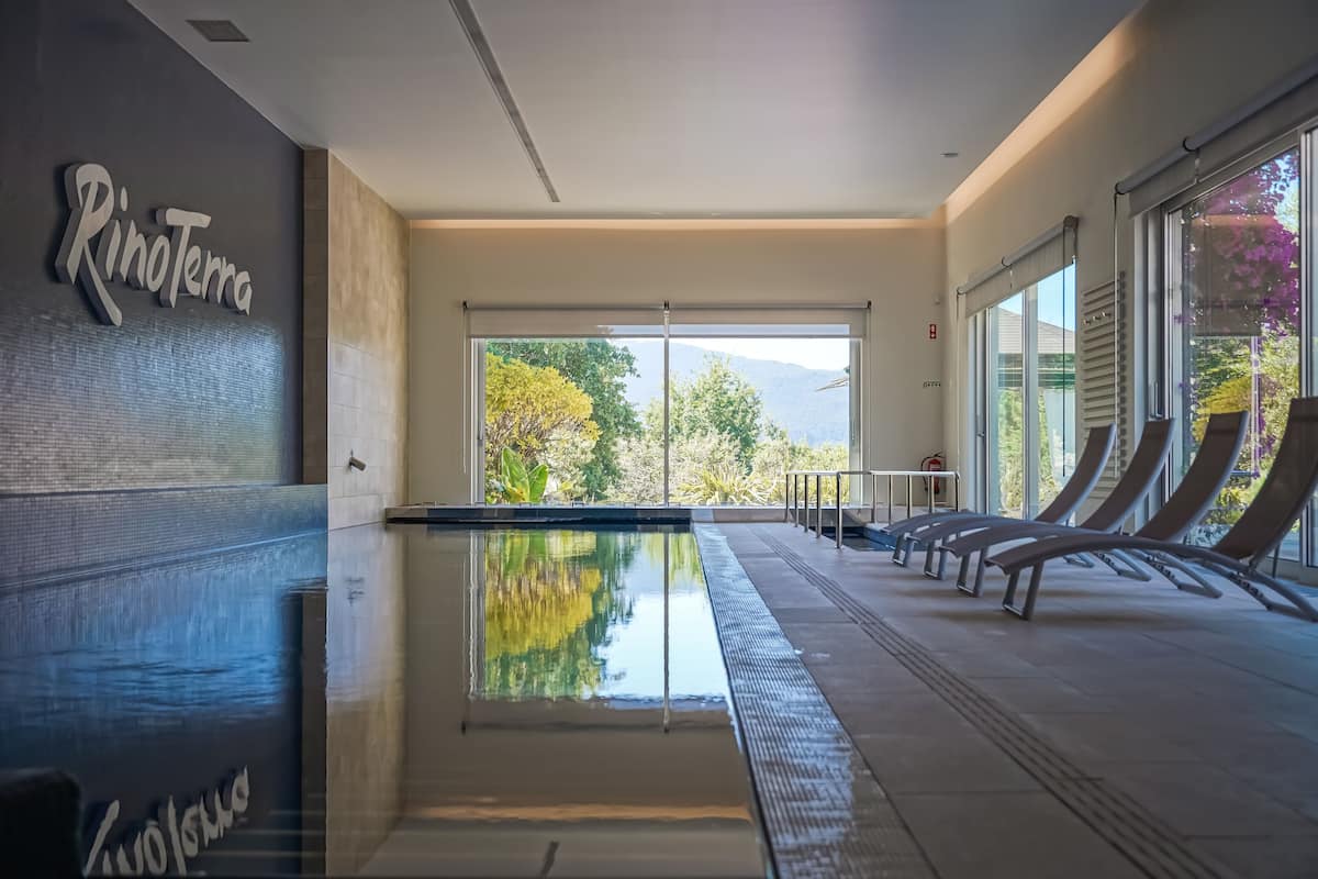Uma área interna moderna com piscina, paredes de vidro transparente, vista para árvores e montanhas, e uma placa com a inscrição 'RinoTerra'.