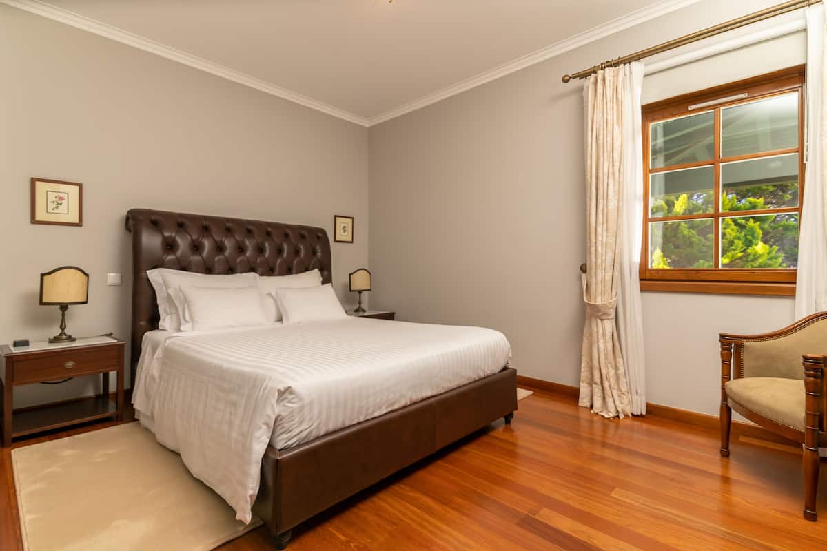 Quarto com uma cama grande, piso de madeira, mesa de cabeceira com luminária, uma cadeira e janela com cortinas.