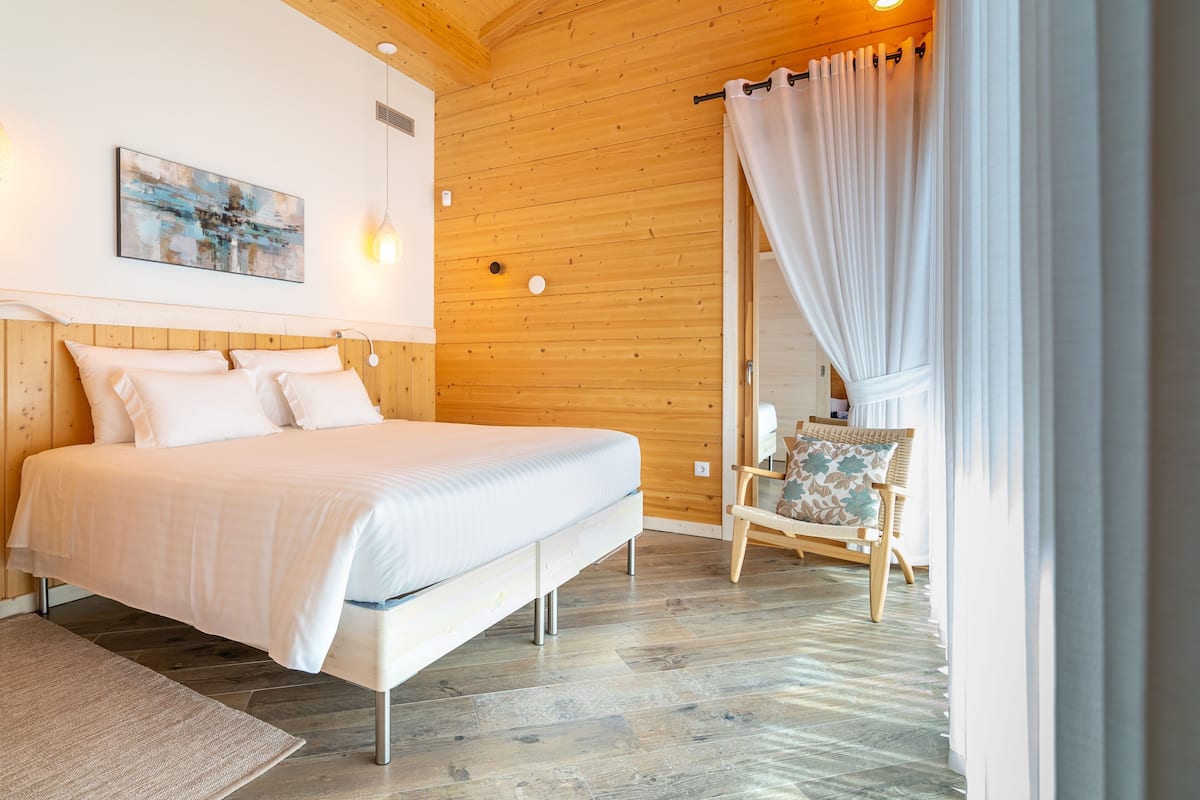 Quarto com teto de madeira, cama com roupa de cama branca, cadeira de vime e uma grande janela com cortinas brancas.