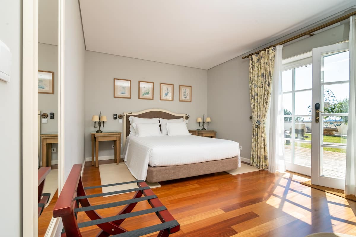 Quarto com cama, mesas de cabeceira, um banco e vista do exterior por uma grande janela.