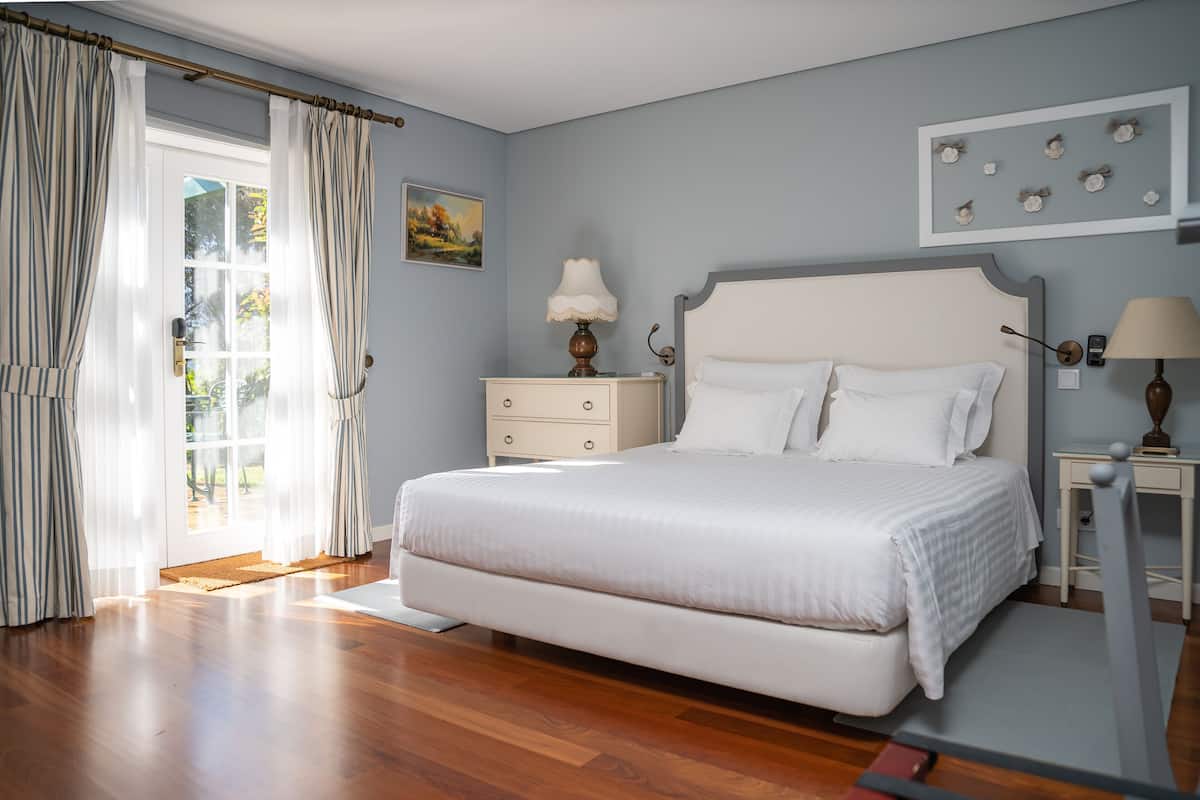 Quarto com cama grande, mesa de cabeceira, abajur e janela com cortinas.