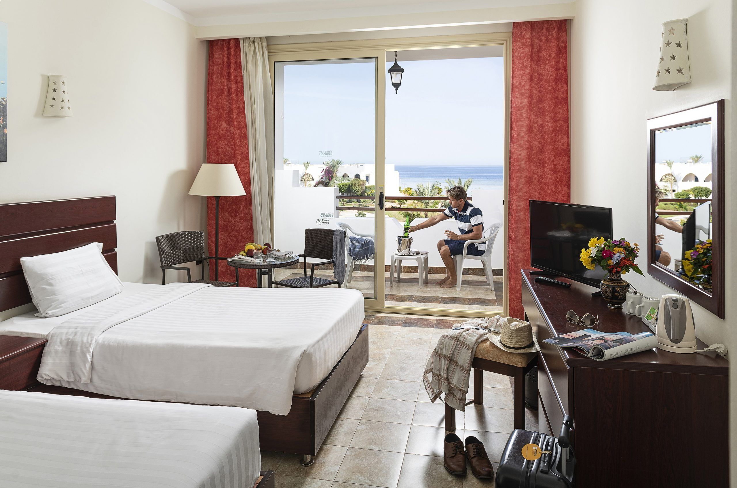 Quarto de hotel com duas camas, televisão, secretária com luminária, cadeira e vista para o mar.