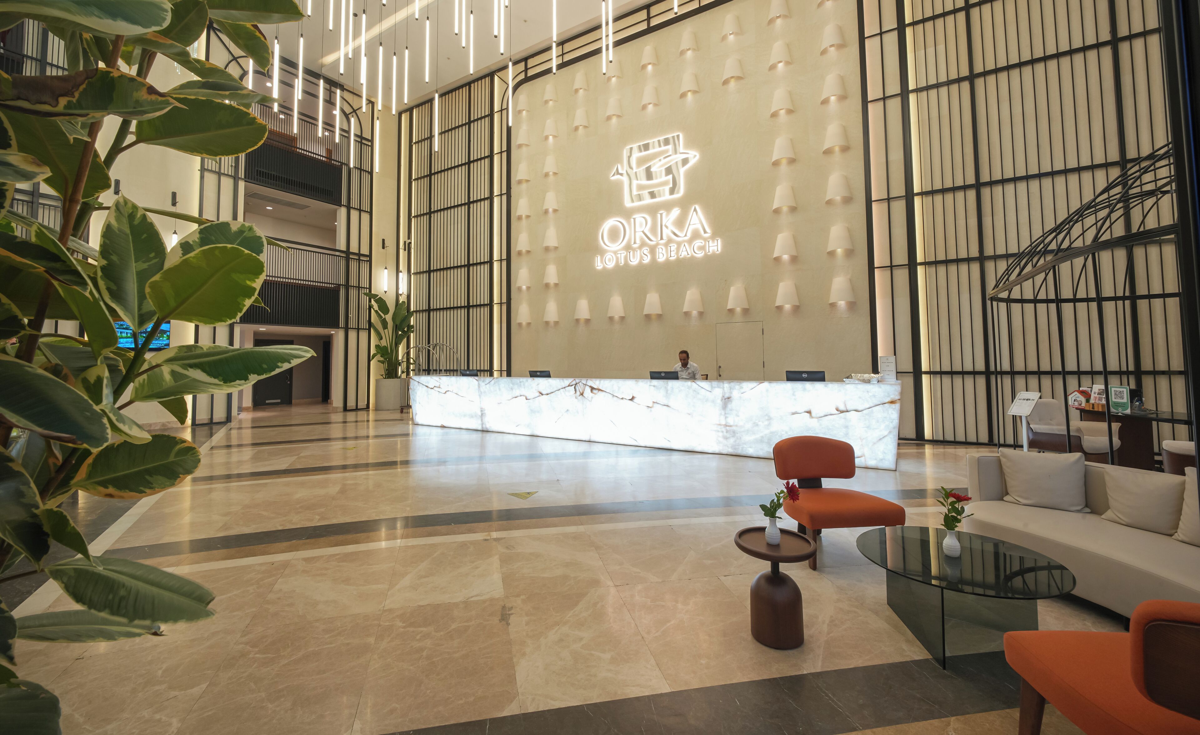 Uma área de recepção moderna com balcão de mármore, um recepcionista e uma área de estar com cadeiras laranja e uma mesa de centro de vidro.