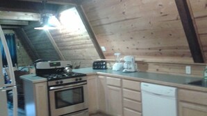 Cozinha equipada com fogão a gás, forno e máquina de lavar loiça. Há uma janela com cortinas e um ventilador de teto.