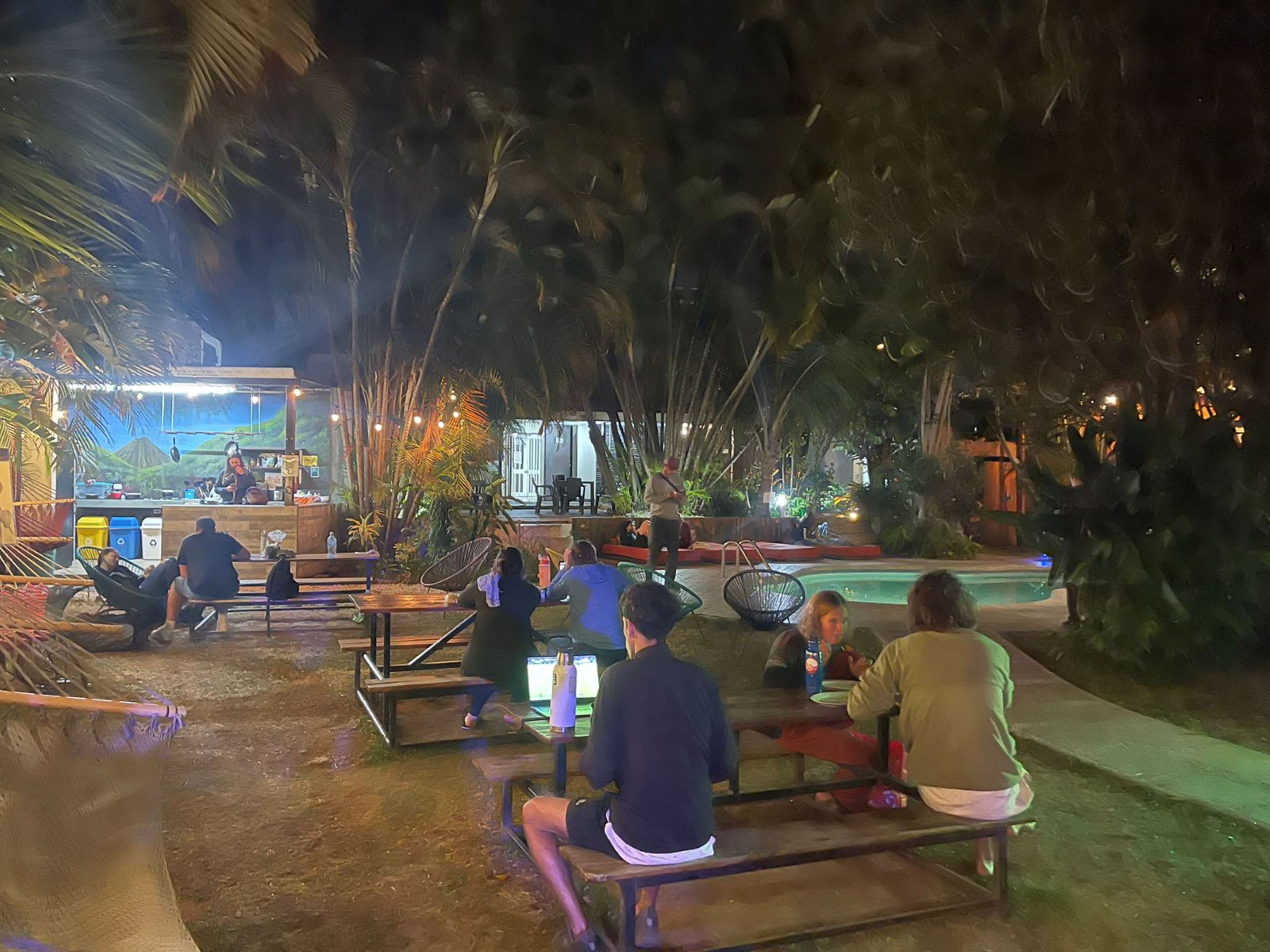Área de refeições ao ar livre com mesas e cadeiras, rodeada por plantas tropicais e uma piscina.
