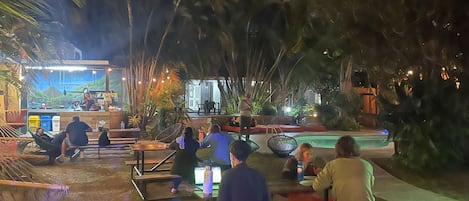 Área de refeições ao ar livre com mesas e cadeiras, rodeada por plantas tropicais e uma piscina.