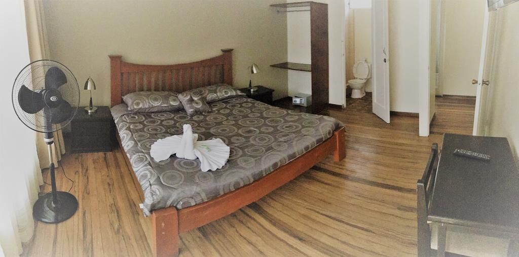 Quarto com cama de madeira, ventilador ao lado da cama, mesa de cabeceira, luminária e porta do banheiro.