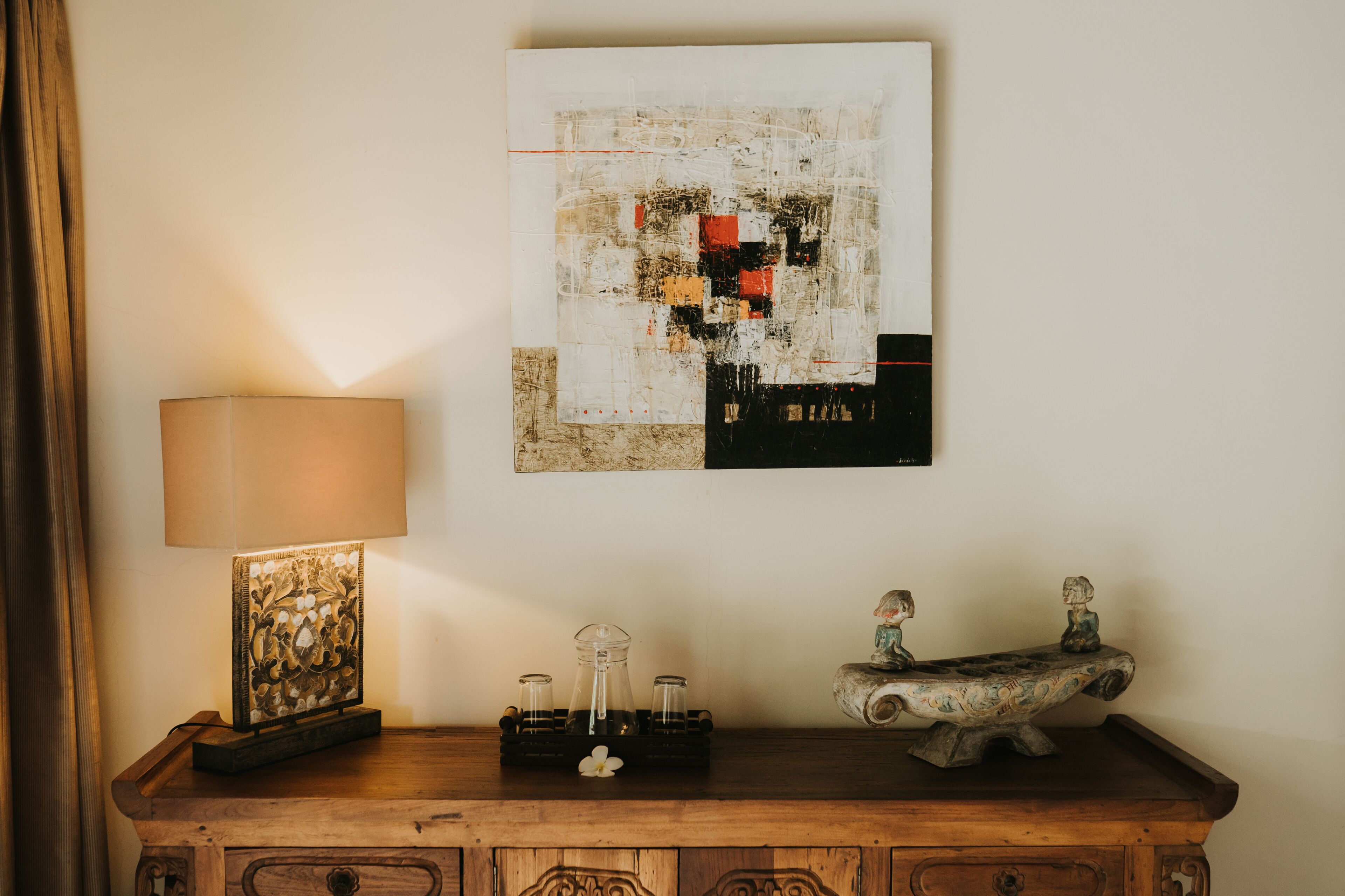 Uma cômoda de madeira com uma luminária, um objeto decorativo e uma obra de arte abstrata emoldurada.