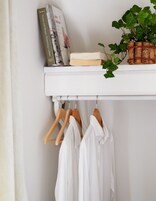 Estante branca com livros e um vaso de planta. Abaixo, camisas brancas penduradas em cabides de madeira.