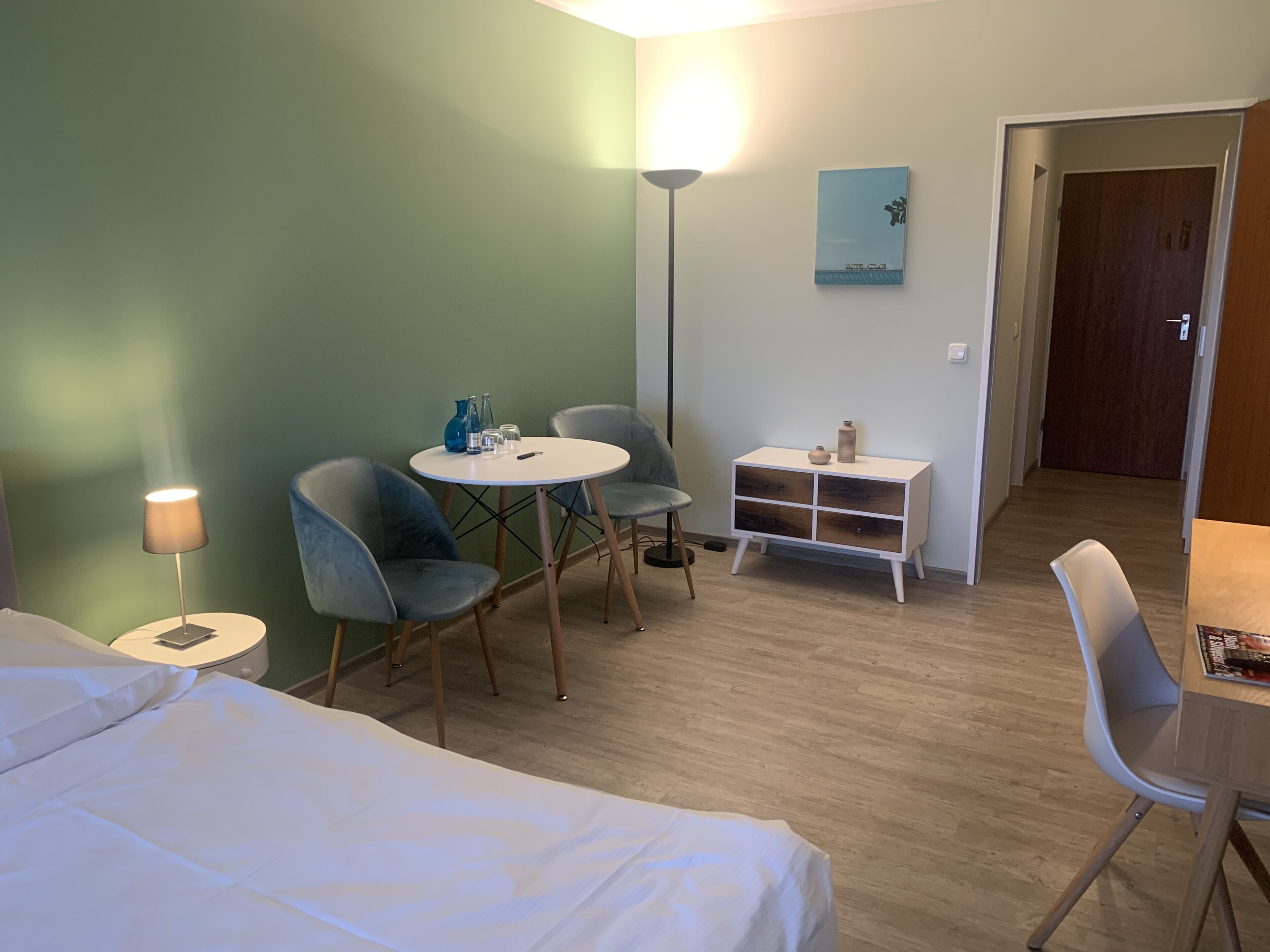 Quarto de hotel com cama, mesa redonda com cadeiras, armário branco pequeno e abajur.