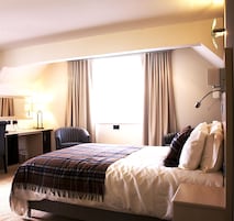 Quarto de hotel com cama, escrivaninha, cadeira e janela com cortinas.