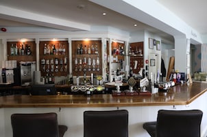Uma área de bar com várias prateleiras de garrafas de bebidas, um balcão com um caixa registradora e banquetas.