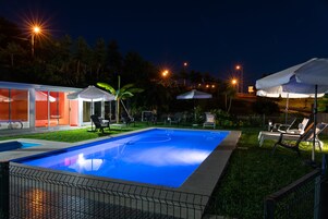 Piscina com superfície de água azul cristalina, rodeada por uma área cercada. Há espreguiçadeiras e guarda-sóis ao redor da piscina.