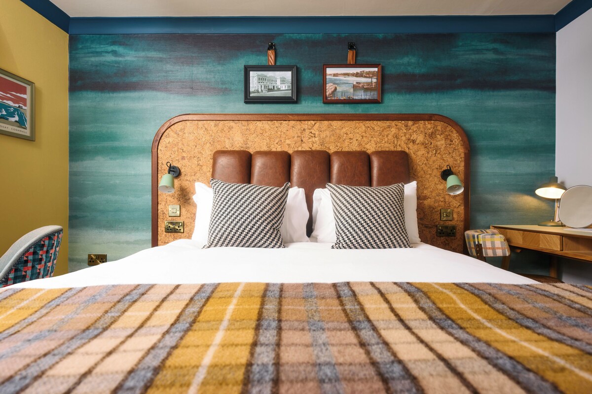 Quarto de hotel com uma cama grande, cabeceira de madeira, duas imagens emolduradas na parede, uma escrivaninha com luminária e uma cadeira.