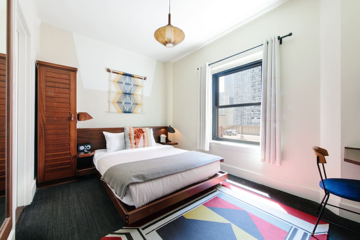 Quarto de hotel moderno com uma cama grande, uma janela com cortinas, uma escrivaninha com cadeira e vista para a cidade.