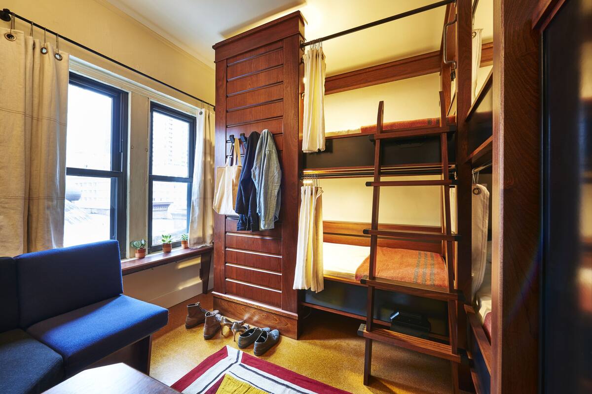Quarto compacto com beliche, sofá, uma pequena mesa e janela com cortinas.