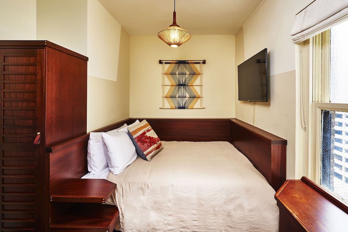 Quarto com cama de madeira, mesa de cabeceira, guarda-roupa, televisão na parede e decoração geométrica na parede.