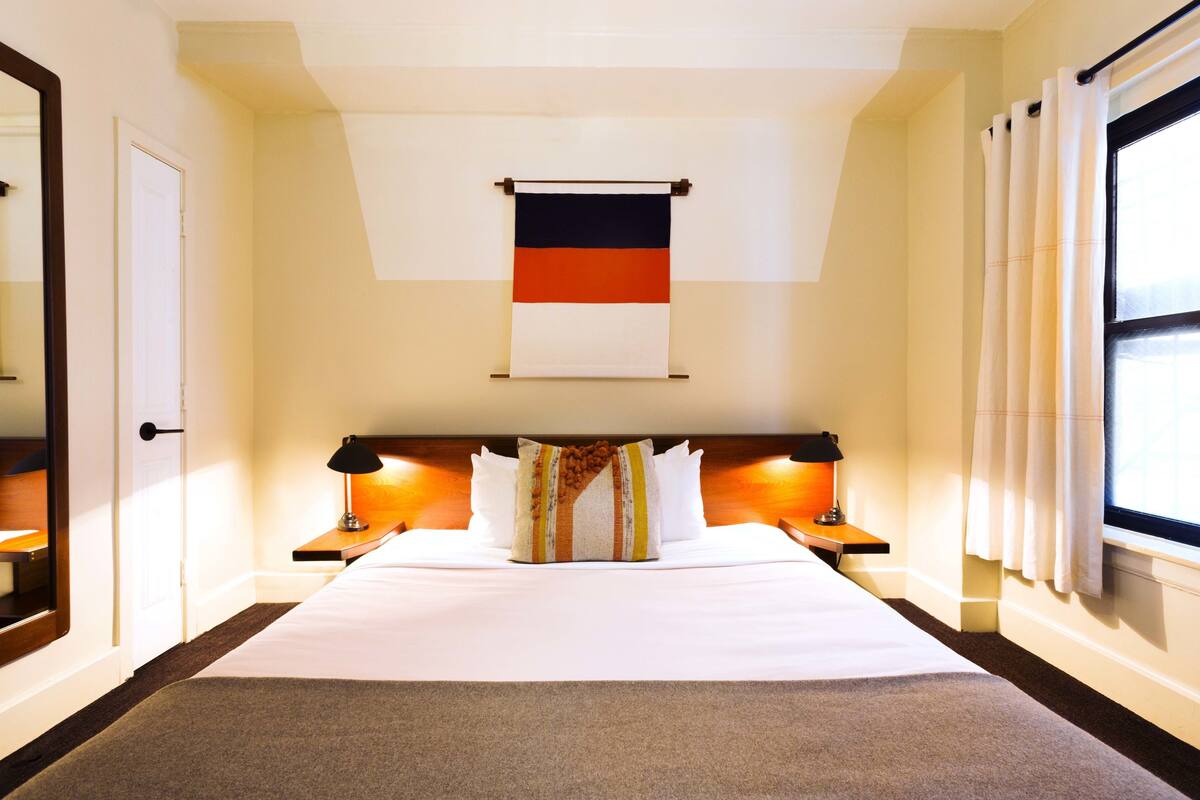 Quarto de hotel com uma cama grande, duas mesas de cabeceira com luminárias, uma bandeira na parede e uma janela com cortinas.
