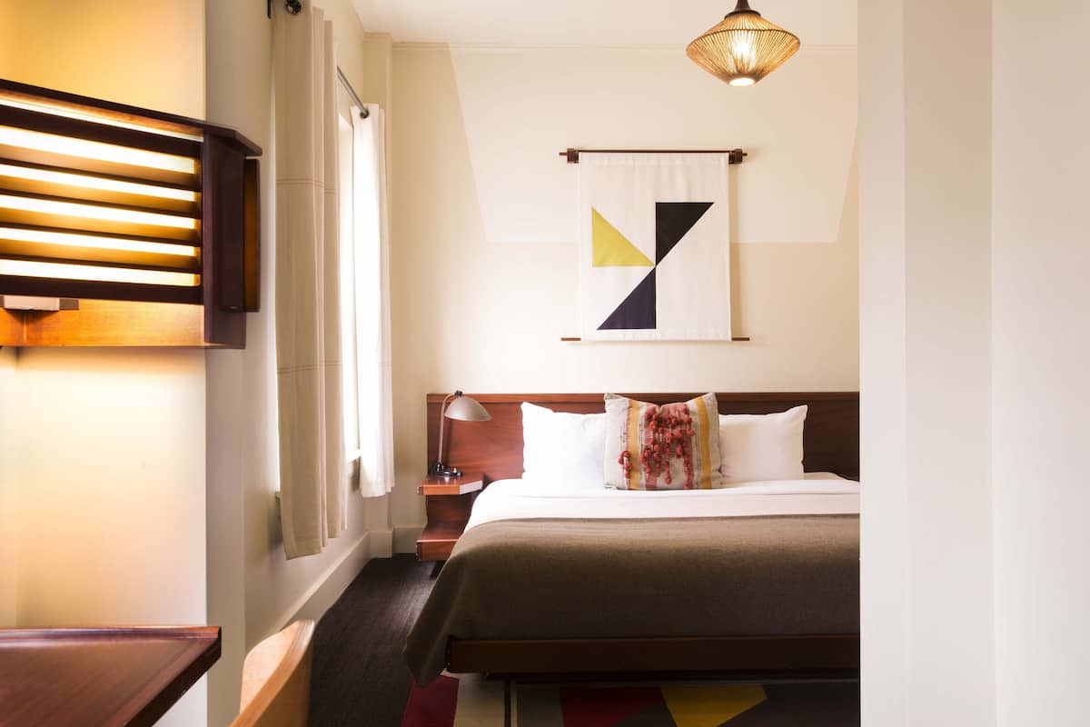 Quarto de hotel com cama, mesa de cabeceira, luminária e um painel de parede geométrico.