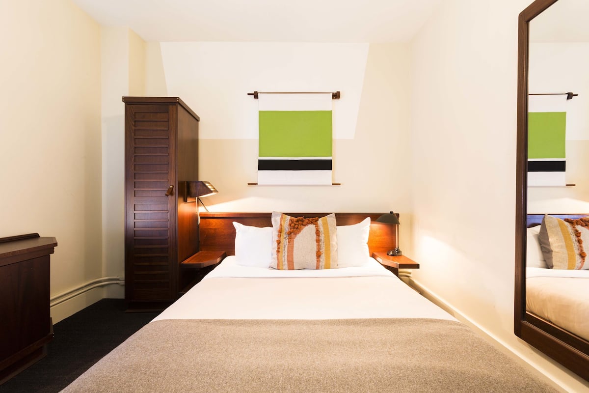 Quarto de hotel com cama, mesa de cabeceira, luminária, guarda-roupa e espelho.