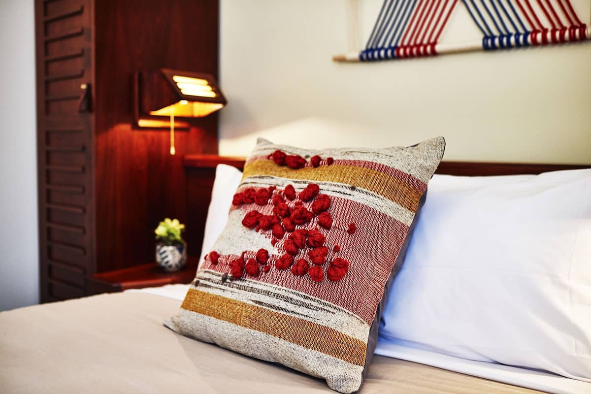 Uma cama com travesseiro estampado com um padrão floral vermelho, uma porta de madeira e uma luminária fixada na parede.