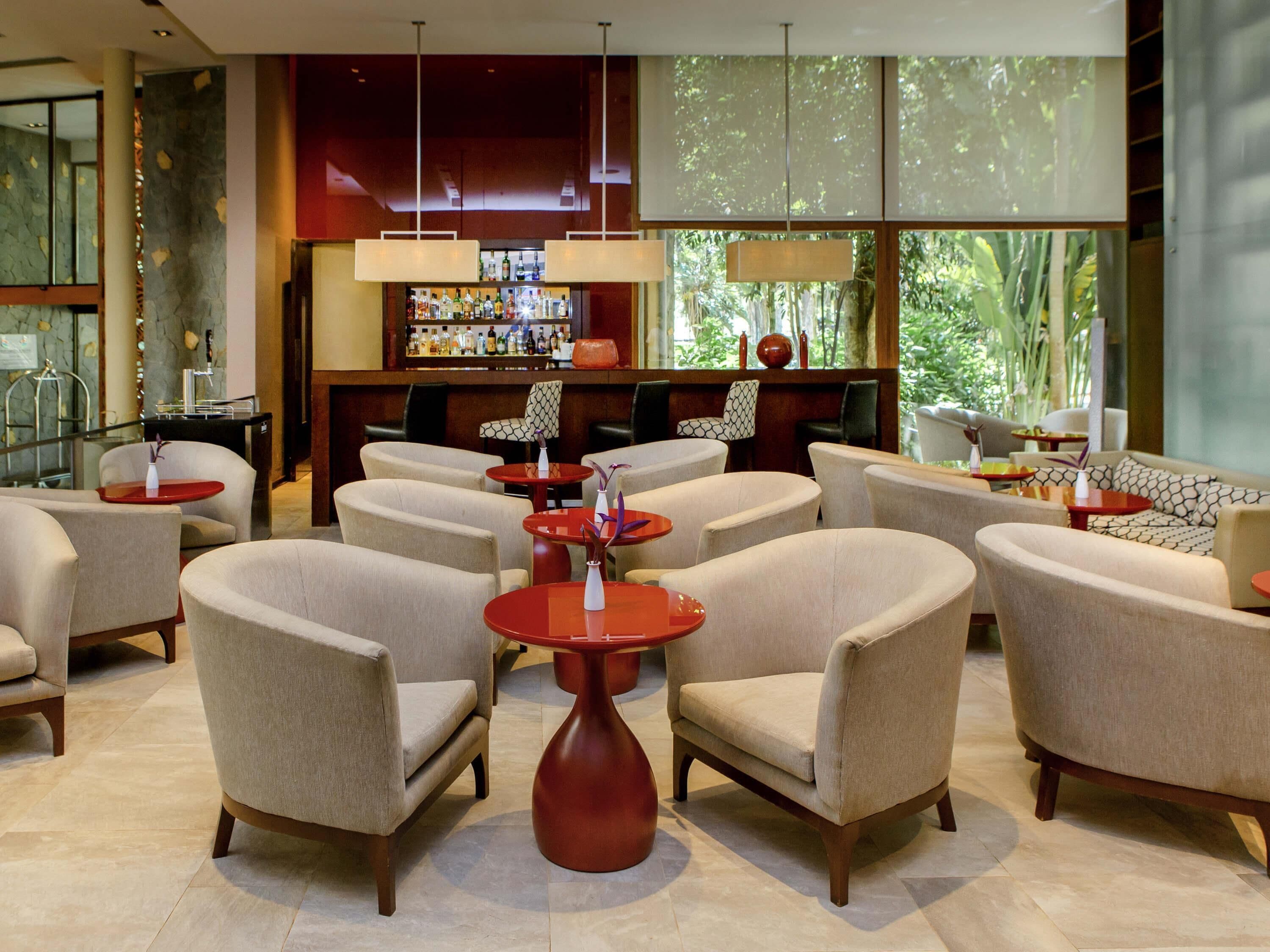 Uma área de estar moderna com sofás e mesas, um bar com garrafas e amplas janelas com vegetação lá fora.