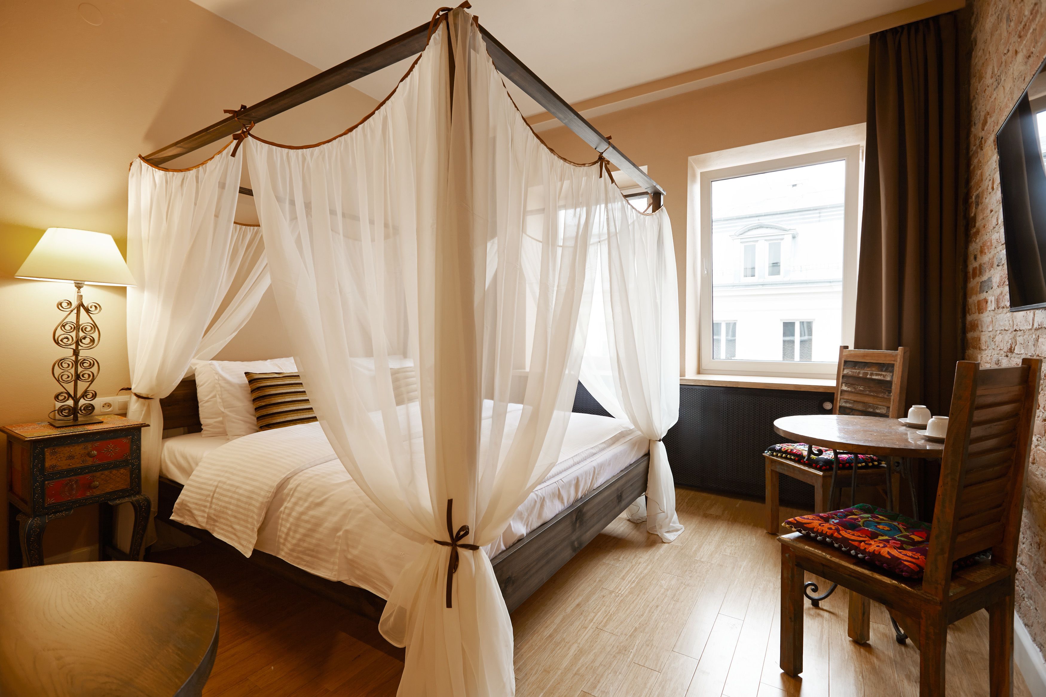 Quarto com cama de dossel, mesa de cabeceira com abajur, uma cadeira e vista para um edifício pela janela.