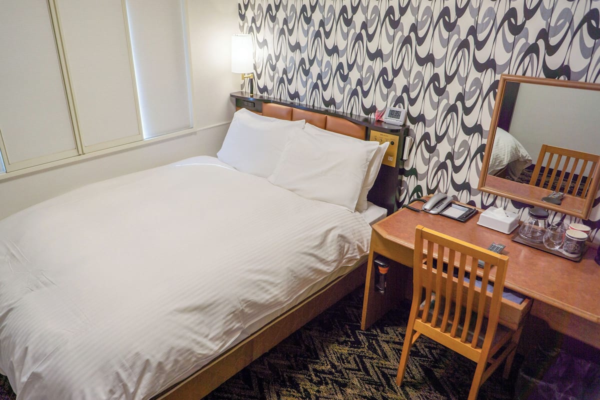 Quarto de hotel com cama, escrivaninha, cadeira, telefone e espelho.