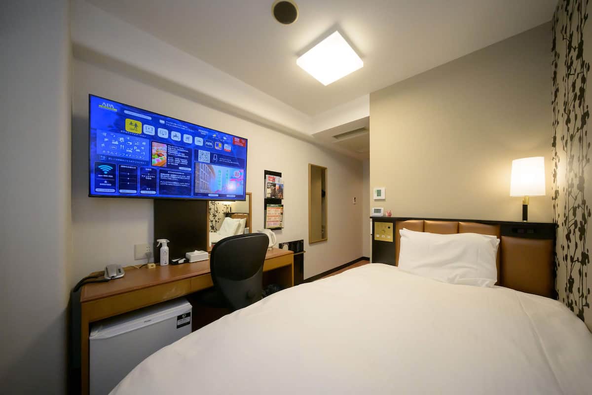 Quarto de hotel com cama, escrivaninha, monitor de computador, cadeira, luminária e pequeno frigorífico.