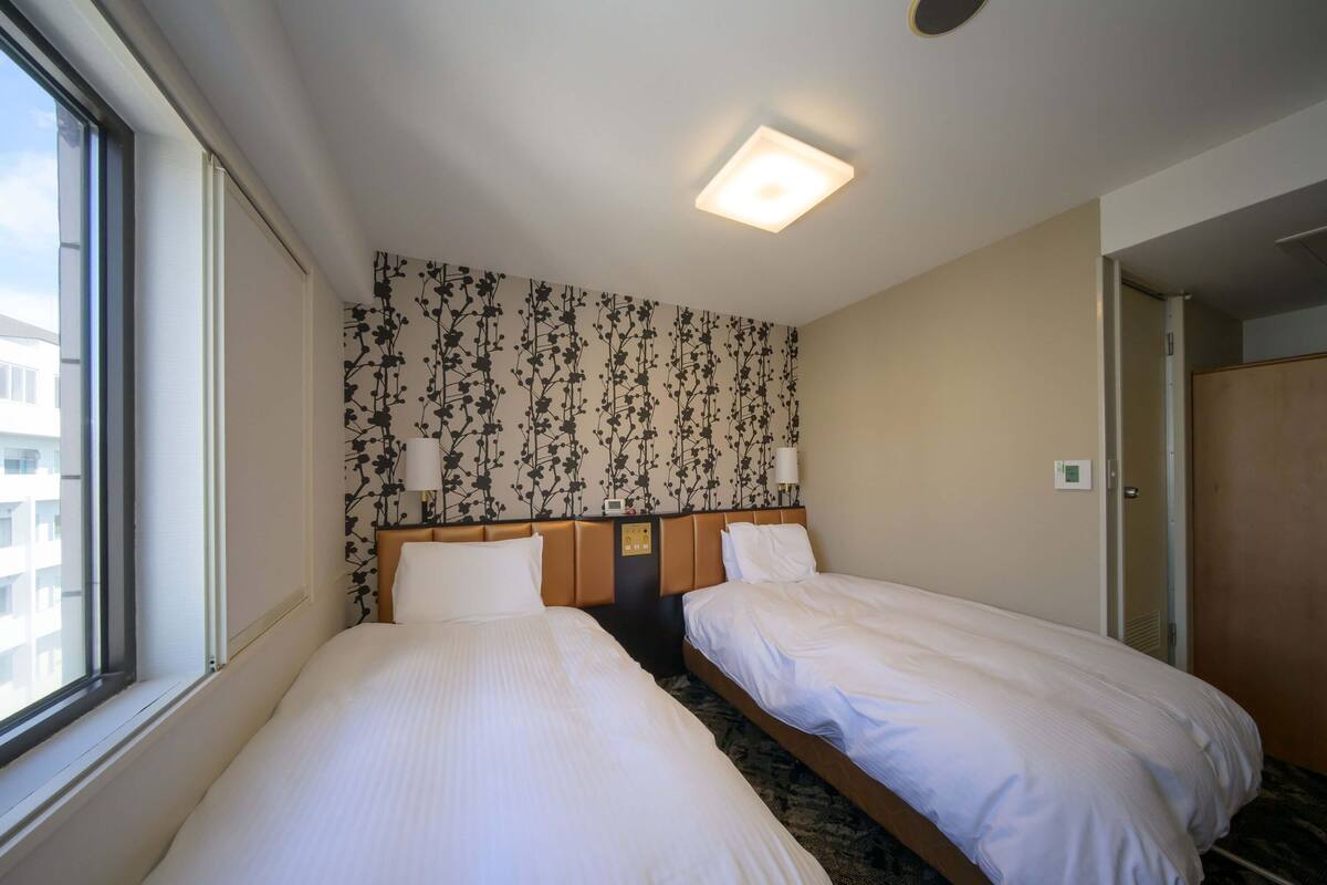 Quarto de hotel com duas camas, uma janela, papel de parede floral e uma luminária no teto.