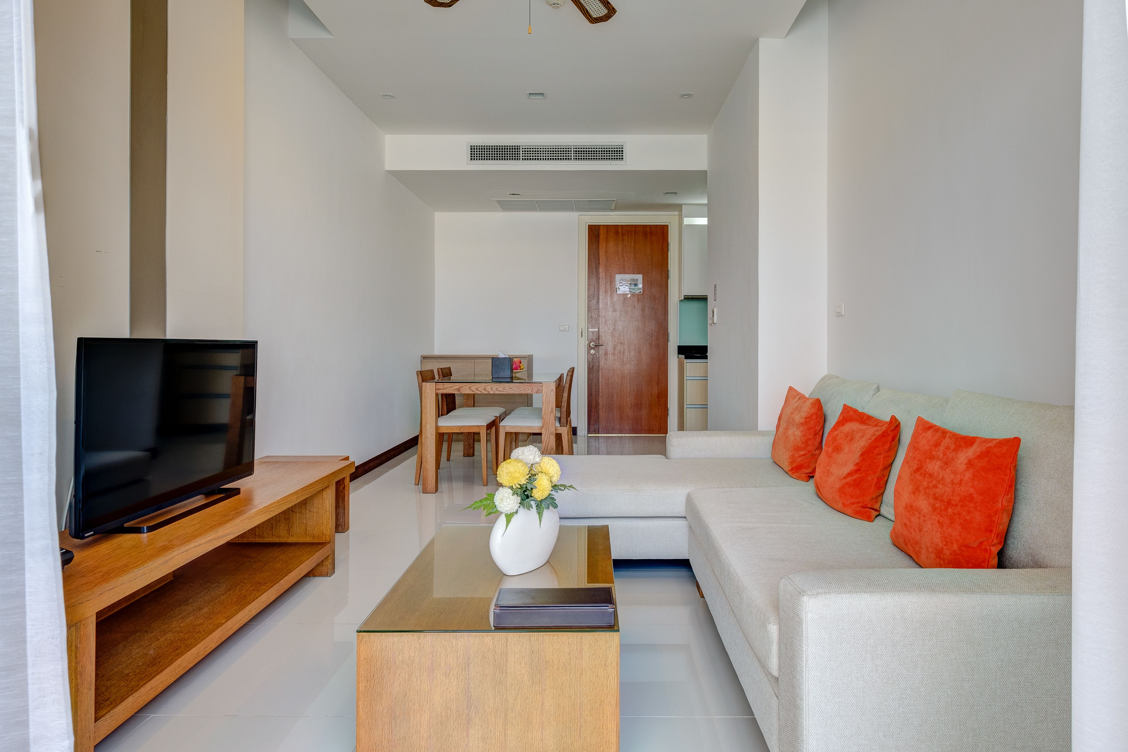 Uma sala de estar moderna com uma TV de tela plana sobre um suporte de madeira, uma mesa de centro de vidro com um vaso de flores, um sofá cinza com almofadas laranja e uma mesa de jantar de madeira com cadeiras.