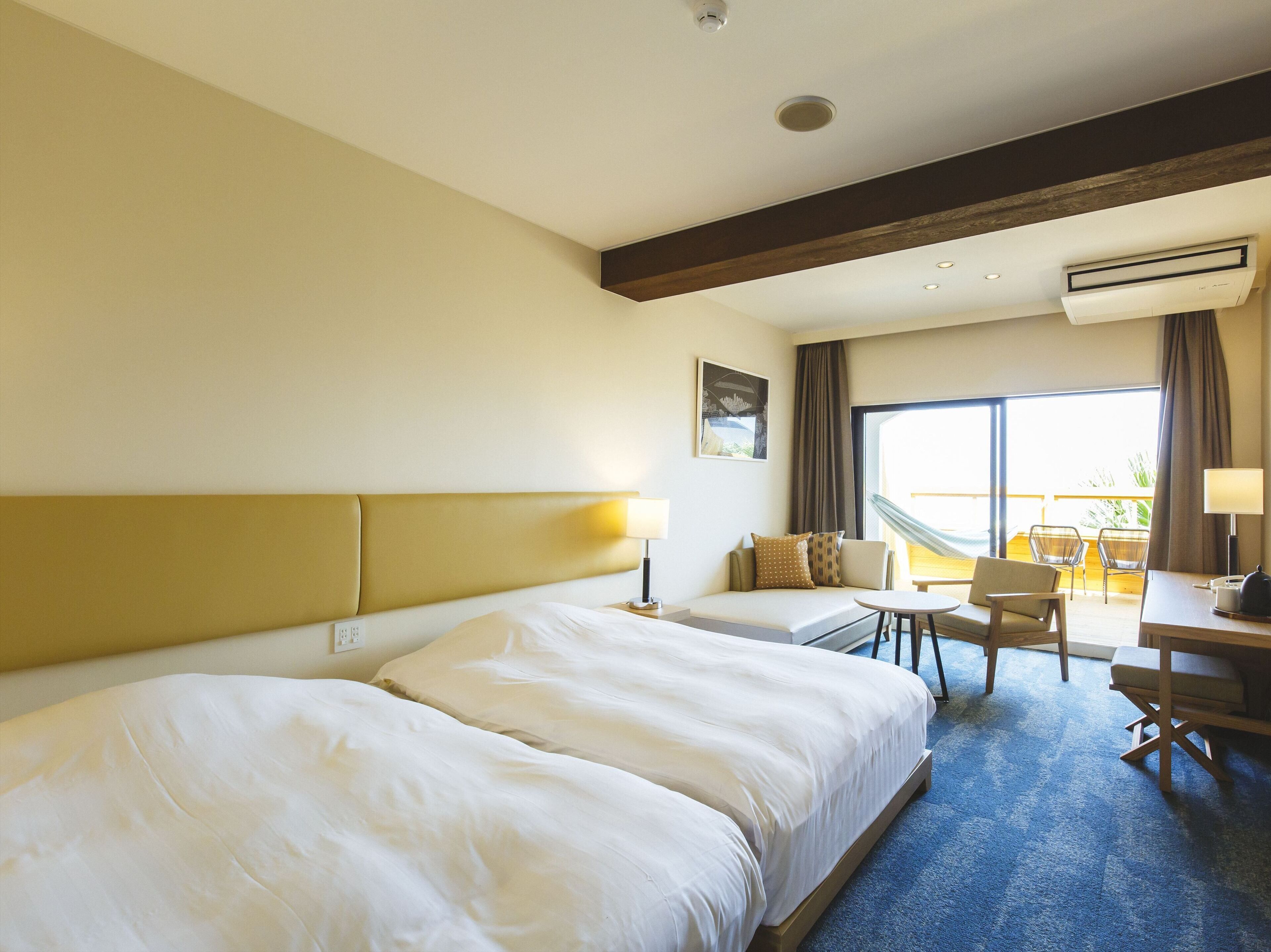 Quarto de hotel com duas camas, uma escrivaninha, uma cadeira e uma varanda com vista.