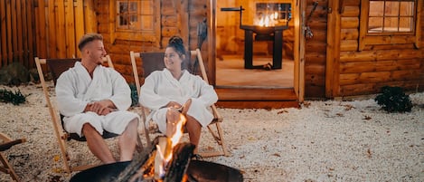 Duas pessoas vestidas de branco sentadas em cadeiras perto de uma lareira a lenha em uma cabana de toras com piso de cascalho.