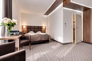 Quarto de hotel moderno com uma cama, mesas de cabeceira, escrivaninha e uma cadeira. Há um vaso com flores na escrivaninha e um abajur na parede.