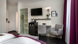 Quarto de hotel com cama, escrivaninha, cadeira, televisão e uma pequena planta.