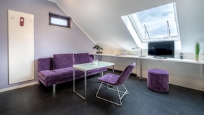 Quarto moderno com sofá roxo, uma mesinha, uma cadeira roxa, um banquinho redondo, uma televisão e um claraboia.