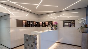 Uma área de recepção moderna com um balcão branco, luminária de teto com design geométrico e prateleira na parede com diversos objetos.