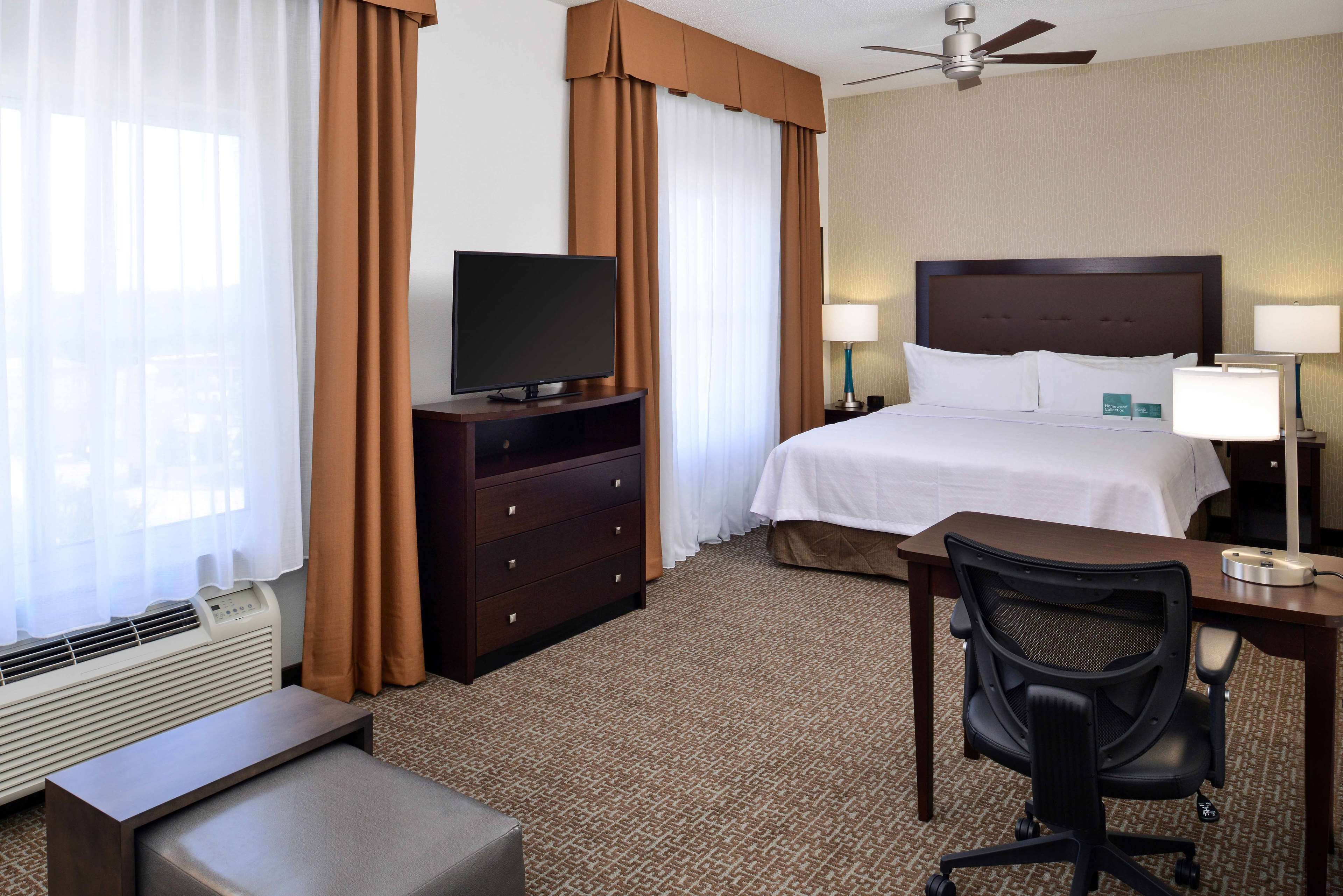 Quarto de hotel com cama, escrivaninha, cadeira, televisão e ventoinha no teto.