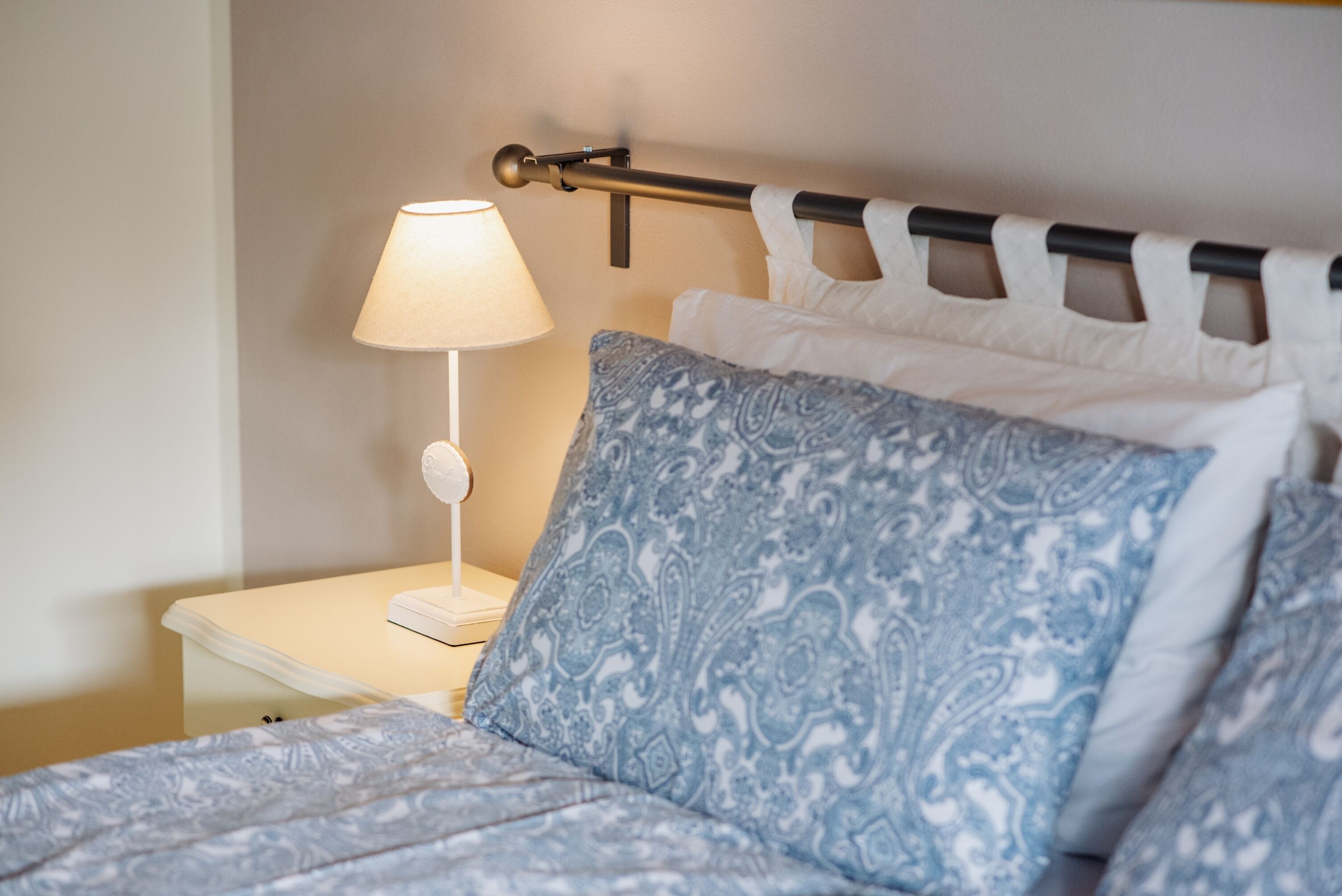 Cama bem arrumada com roupa de cama estampada, luminária de cabeceira e cabeceira com barra para cortina.