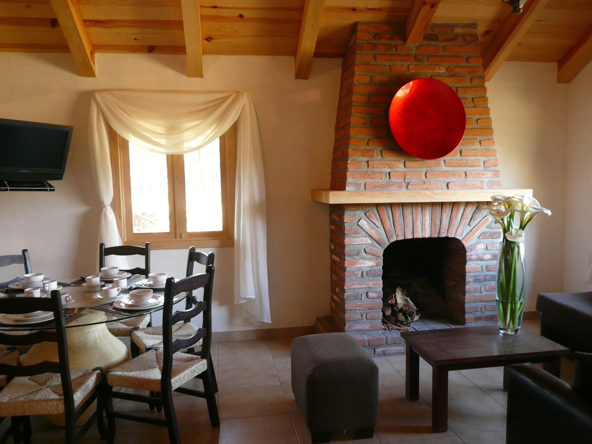Uma sala de estar aconchegante com lareira de tijolos, vigas de madeira, um espaço de refeições com cadeiras e mesa posta para uma refeição, e uma televisão fixada na parede.