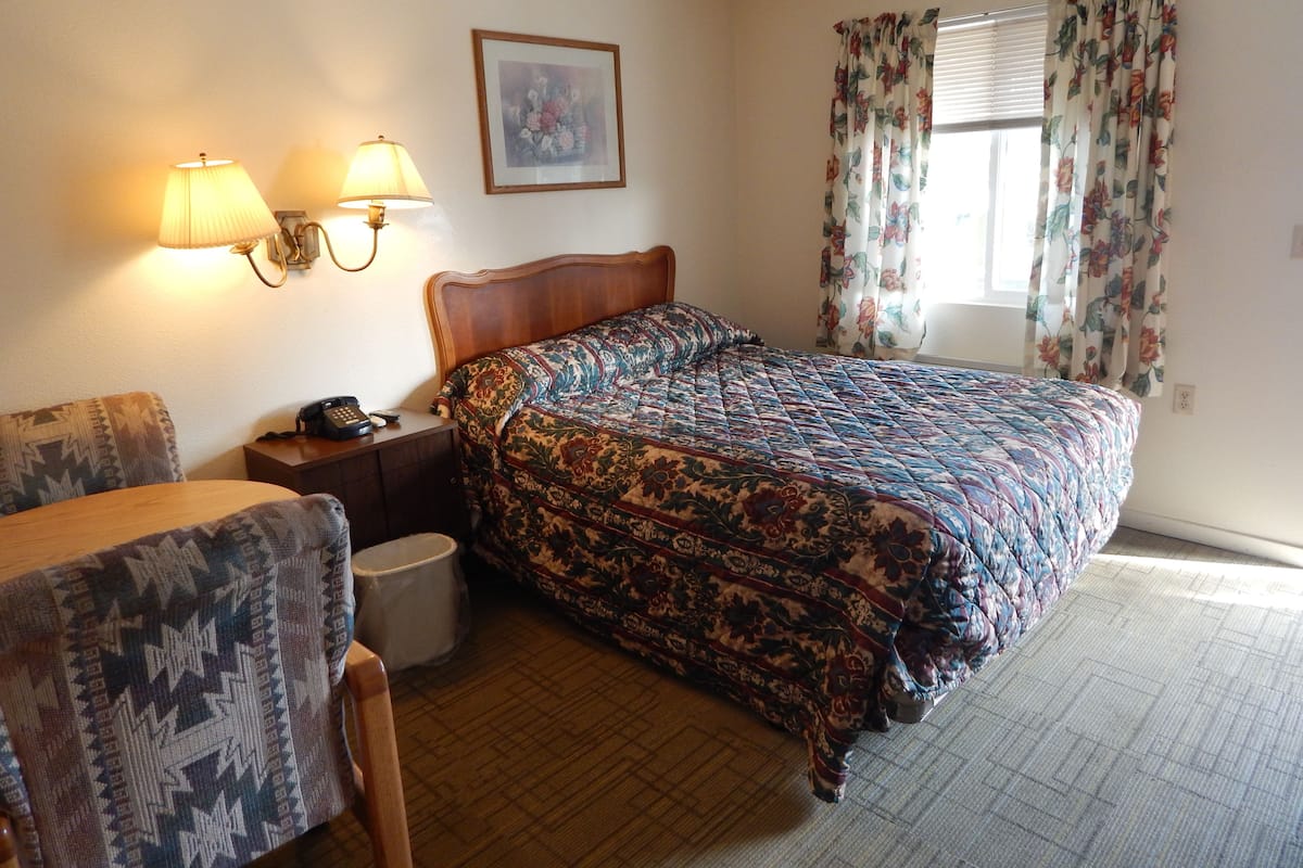 Quarto com uma cama, duas luminárias de cabeceira, um telefone, uma cadeira, uma mesinha e uma janela com cortinas florais.