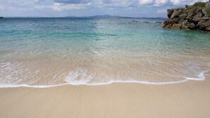 Uma praia com águas turquesa cristalinas, ondas suaves e um afloramento rochoso à direita.