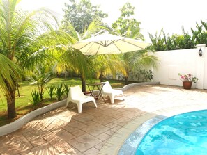Área da piscina com um terraço ladrilhado, duas cadeiras de plástico brancas, um guarda-sol bege e um vaso de planta.