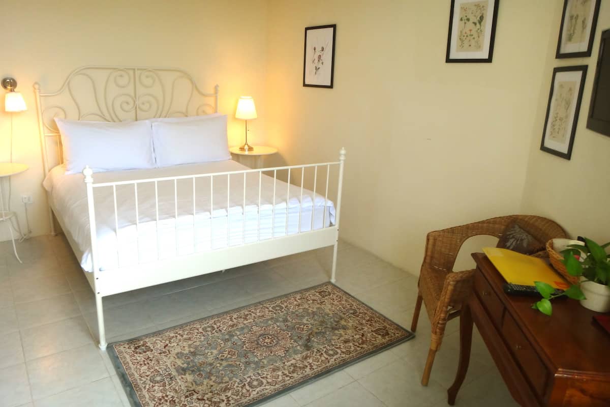 Quarto com cama de metal branca, duas luminárias de cabeceira, tapete estampado, cadeira de madeira e uma mesinha com uma planta.