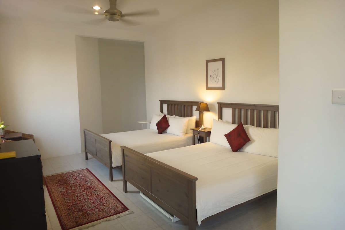 Quarto de hotel com duas camas, um ventilador de teto, uma mesinha com abajur e um quadro na parede.